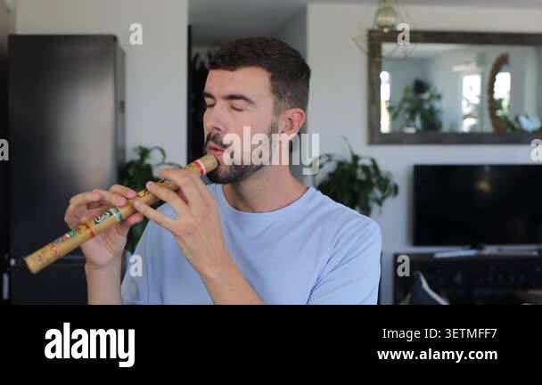 A man playing a wooden flute indoors, with plants and a mirror visible ...
