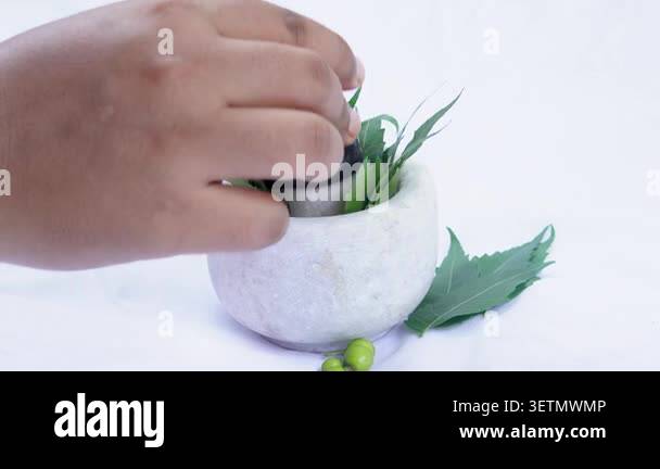 A woman hand holding a pestle and pounds neem leaves in a white stone ...
