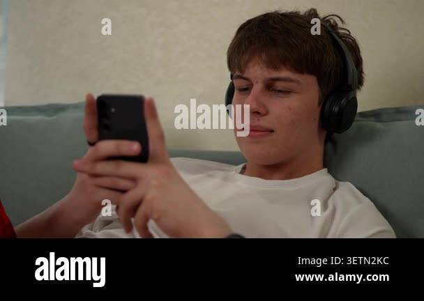 A young boy sits on a couch using his phone and listening to music with ...