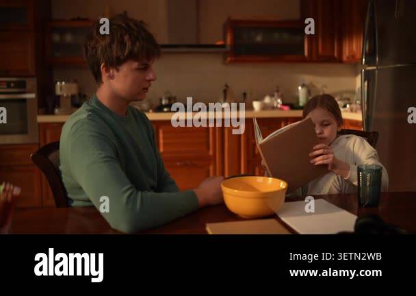 A boy and a girl sit in the kitchen at night. They look at a book while ...