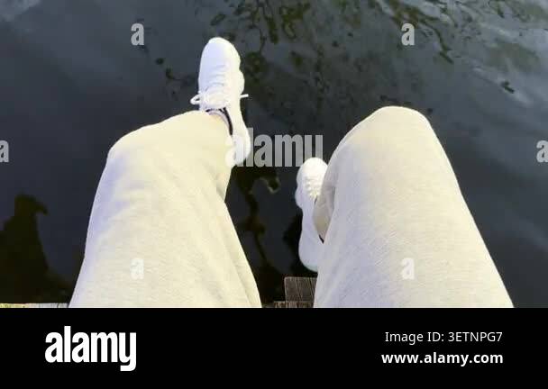 POV of male swinging legs on wood pier above calm lake. Summer sunlight ...