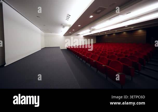 Indoor auditorium with red upholstered seats faces a large white ...