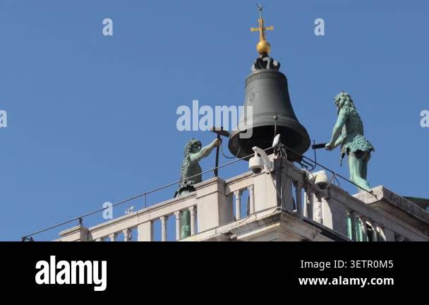 A large bell and bronze figures sit atop a white stone tower over a ...