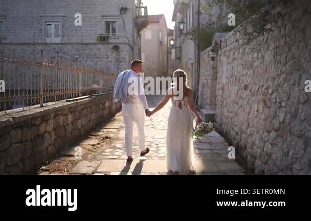 Caucasian couple walking cobblestone alley at golden hour, bride in ...