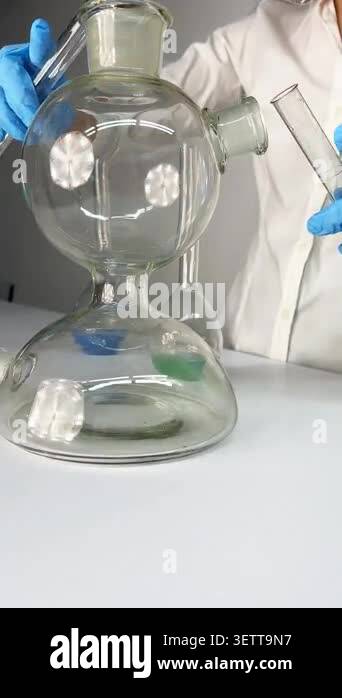 A person in a lab coat mixes clear liquids in a round-bottom flask. The ...