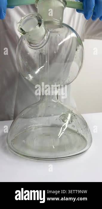 In a lab, a person pours a clear liquid into a glass container. The ...