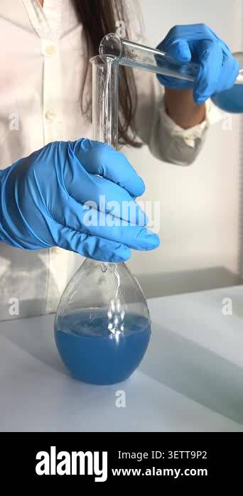 In a laboratory, a person with blue gloves pours a blue liquid from one ...