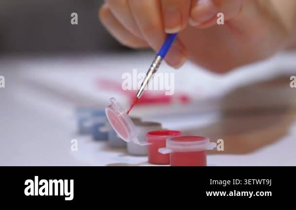Close-up of a hand dipping a thin brush into a small container of red ...