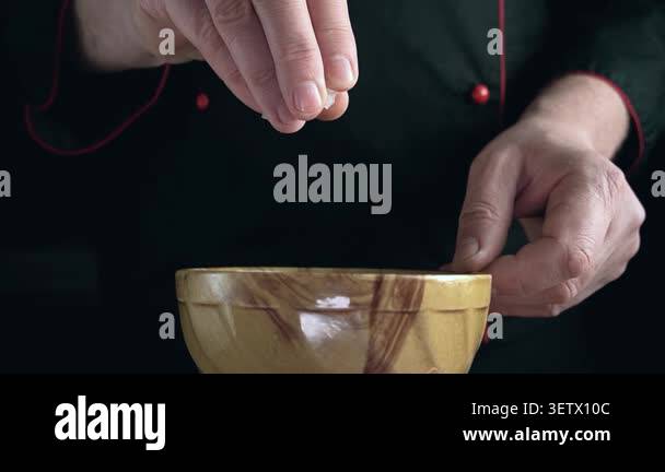 A chef in a black uniform carefully adds salt to a clay bowl ...