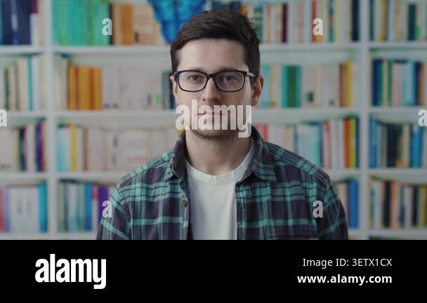 A man who is wearing glasses is standing in front of a tall bookshelf ...