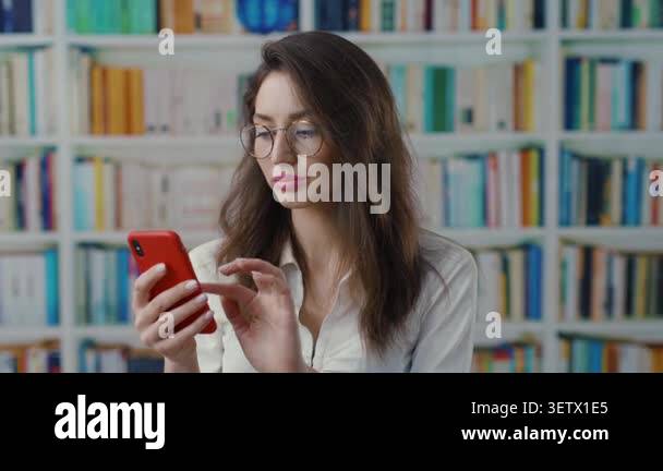 A woman in a library focuses on her red cell phone, blending ...