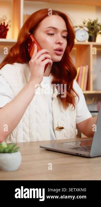 Young woman on home table talks on smartphone while working on laptop ...