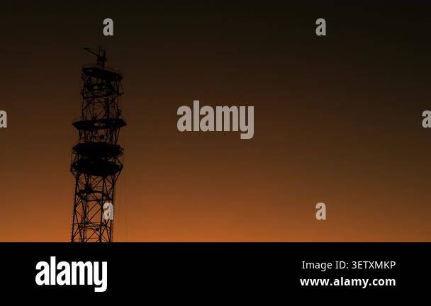 Silhouette of telecommunication tower against glowing orange twilight ...