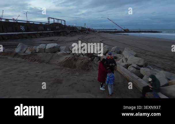 A family stands on a sandy beach near a rocky breakwater, with a crane ...