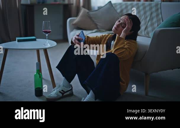Stressed woman watching mobile phone sitting apartment floor with wine ...
