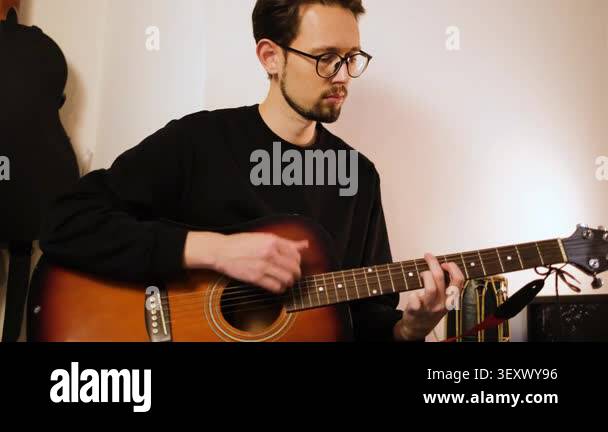 Young brunette musician in glasses playing guitar in his music studio ...