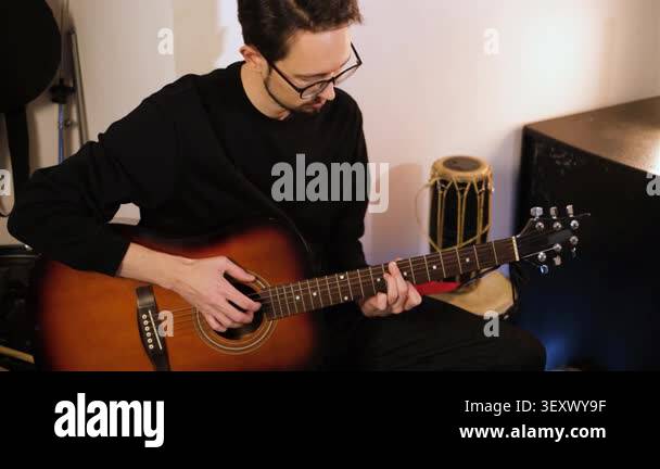 Young brunette musician in glasses playing guitar in his music studio ...