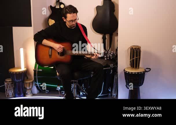 Young brunette musician in glasses playing guitar in his music studio ...