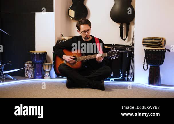 Young brunette musician in glasses playing guitar in his music studio ...