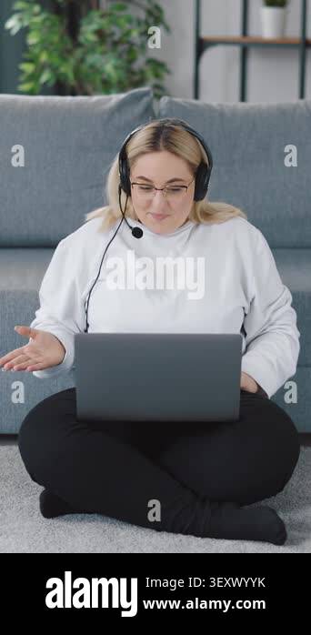A woman sits on a rug using a laptop and wears a headset while focused ...