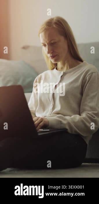 Woman sits on couch with laptop typing and smiling in a cozy home ...