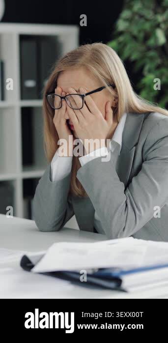 A woman sits at a desk with documents, showing signs of exhaustion and ...