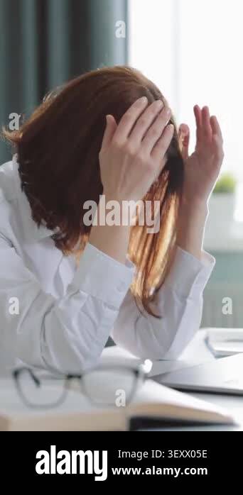 A tired woman sits at a desk with a laptop, hands in her hair, focused ...