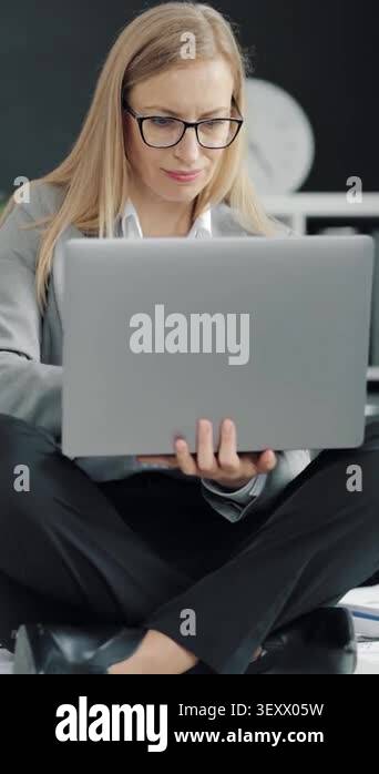 A woman sits on the floor in an office and works on a laptop with focus ...