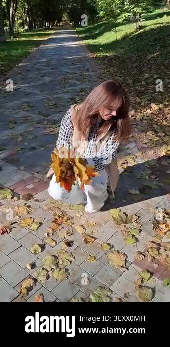 A person kneels on a path in a park, gathering leaves from the ground ...