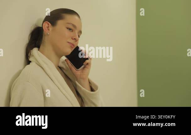 Young woman in a bathrobe leaning against a wall while talking on the ...