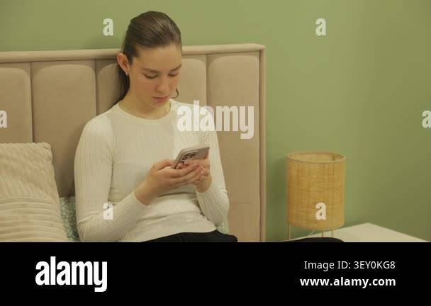 Young woman sitting on her bed, completely absorbed while messaging on ...