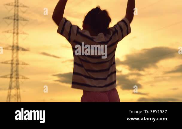 Back view of a happy young girl jumping and raising hands at sunset ...