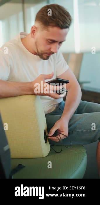 Young man in airport waiting area connects smartphone to charging port ...