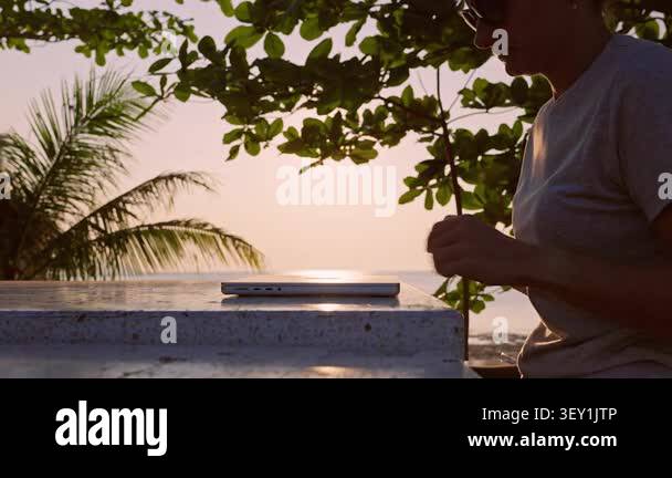 Side View of Woman Opening Laptop at Sunset by Ocean with Tropical ...