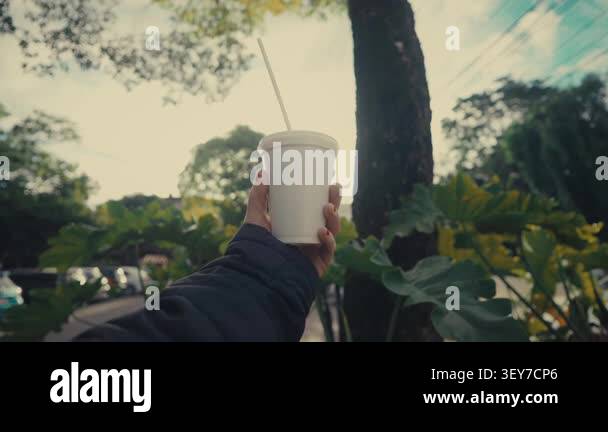 Hand holds a white disposable cup with a straw. Trees and greenery ...
