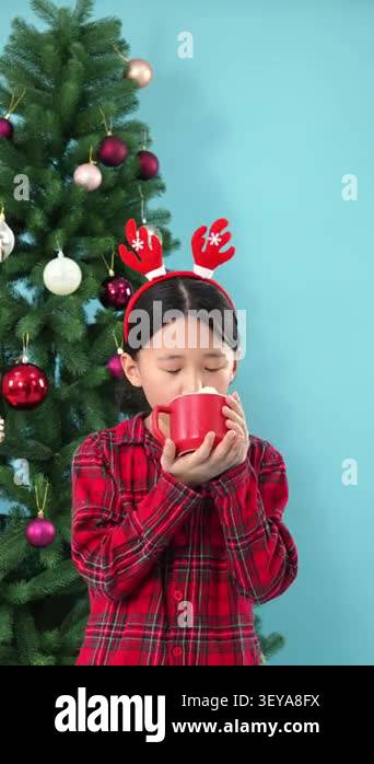 A young girl wearing a holiday headband blows on and sips a warm drink ...