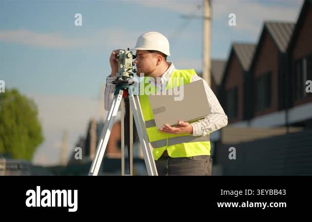 Professional land surveyor in a hard hat and safety vest working with a ...