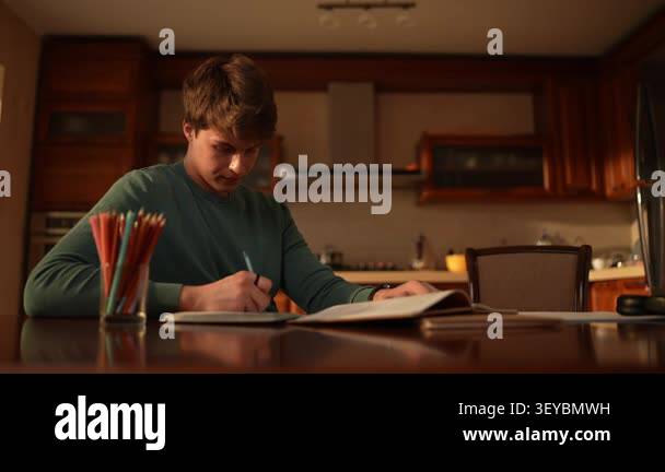 A young man sits at a table in his kitchen, drawing and writing in a ...