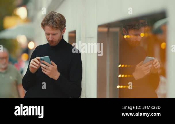 A man stands outside a building looking at his smartphone. He is ...
