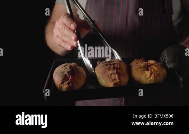 Baker in a striped apron lifts a warm bread roll from a tray using ...