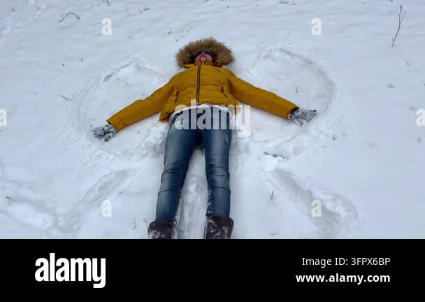 Cheerful woman lying on snowy lawn at forest and making angel. Young ...