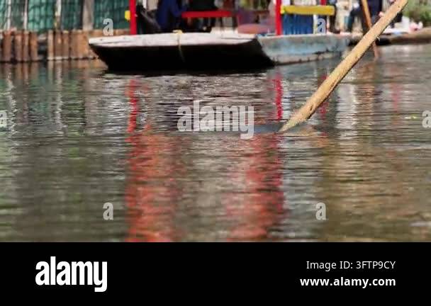 A wooden oar stirs water while moving a brightly colored trajinera boat ...