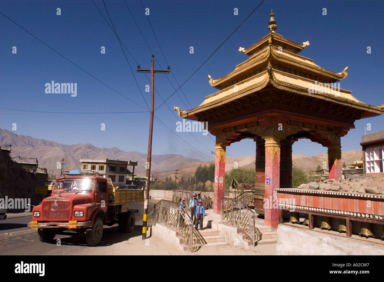 A red Indian truck passing raditional big prayer wheel and prayer wheel ...