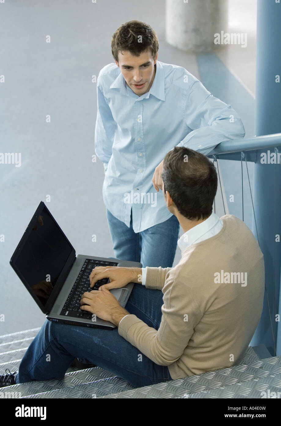 Man sitting on steps, using laptop, speaking to second man Stock Photo ...