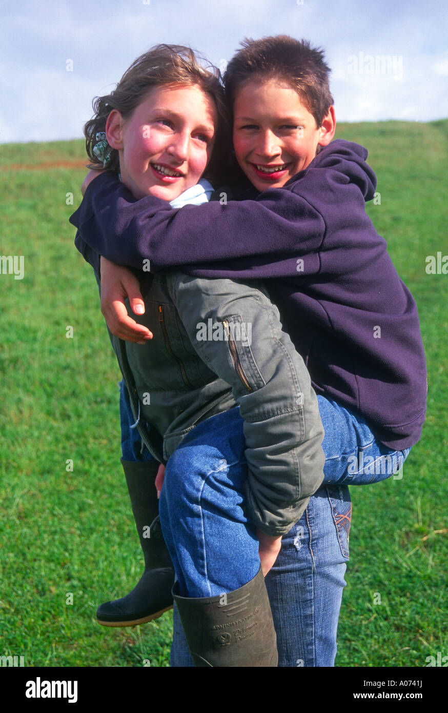 Young girl giving her twin brother a piggyback across a green field ...