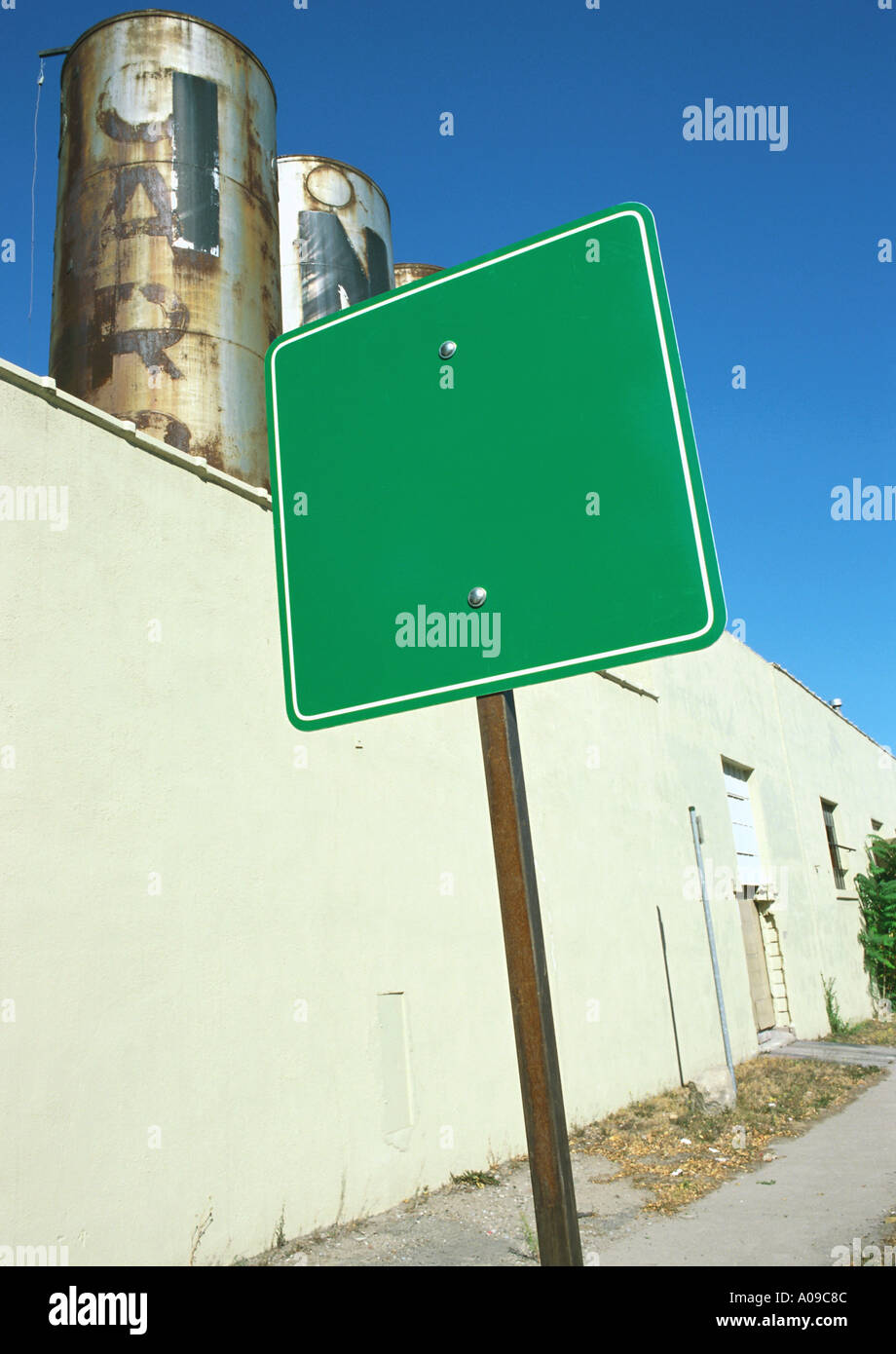 Blank green sign on a rusty metal post with factory storage tanks in ...