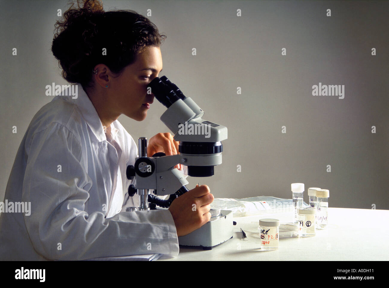 Profile of young woman in white lab coat looking into microscope Stock ...