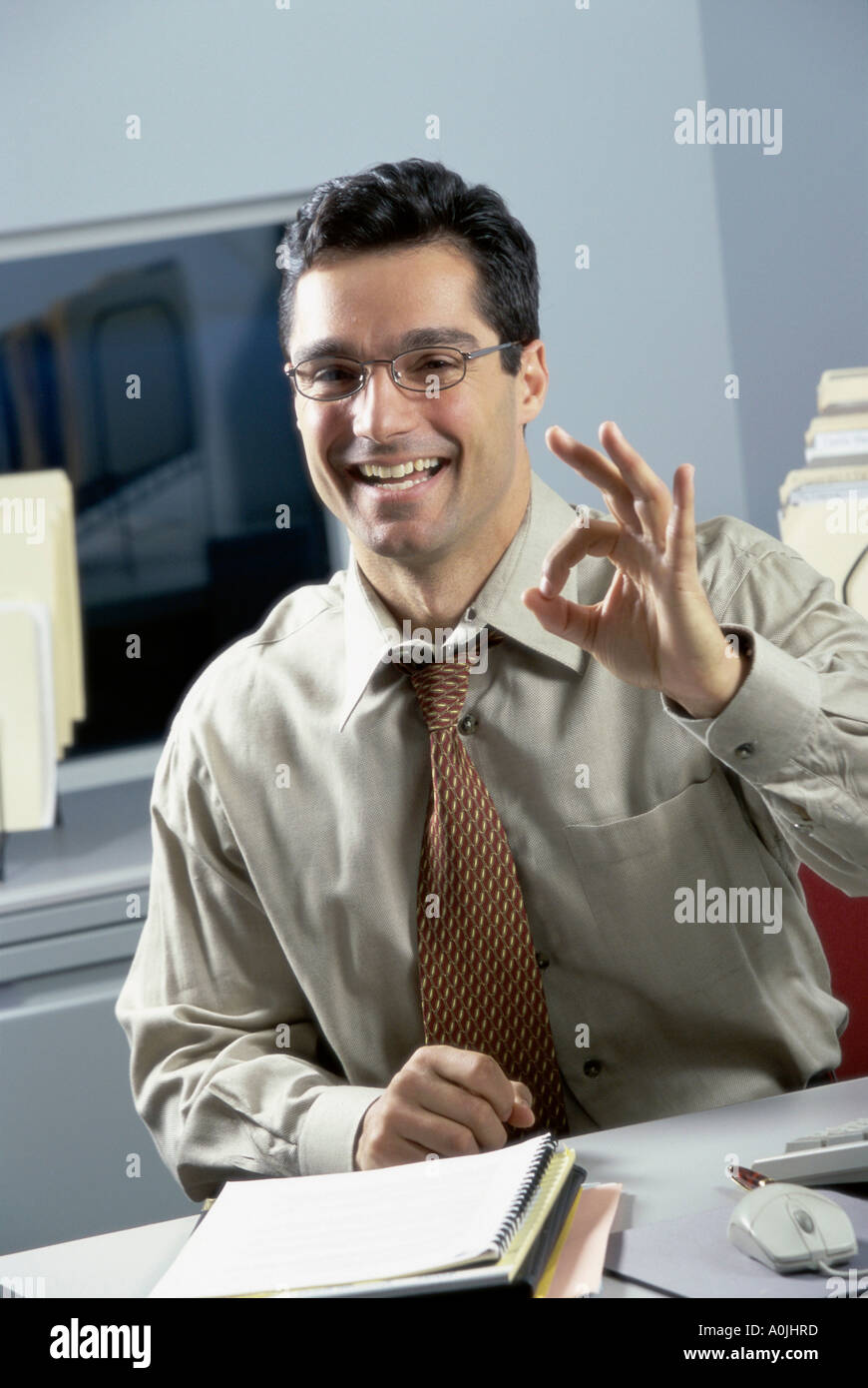 Portrait of a businessman seated at an office desk making an OK sign ...