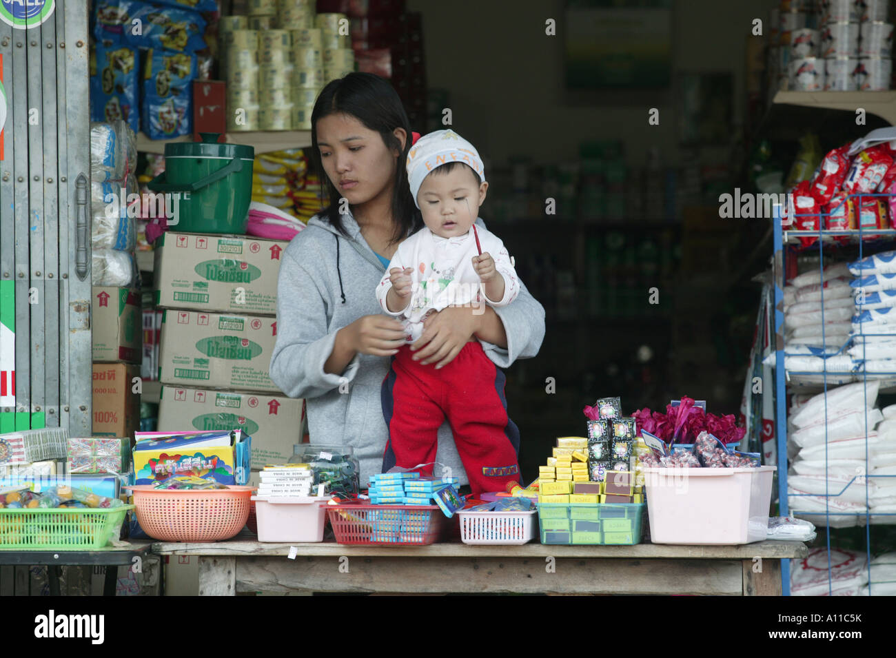 Shop trader holding a baby standing behind table displayed with things ...