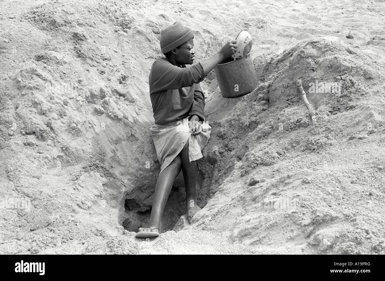 B/W of a woman scooping out water from a dug out hole in a dry river ...